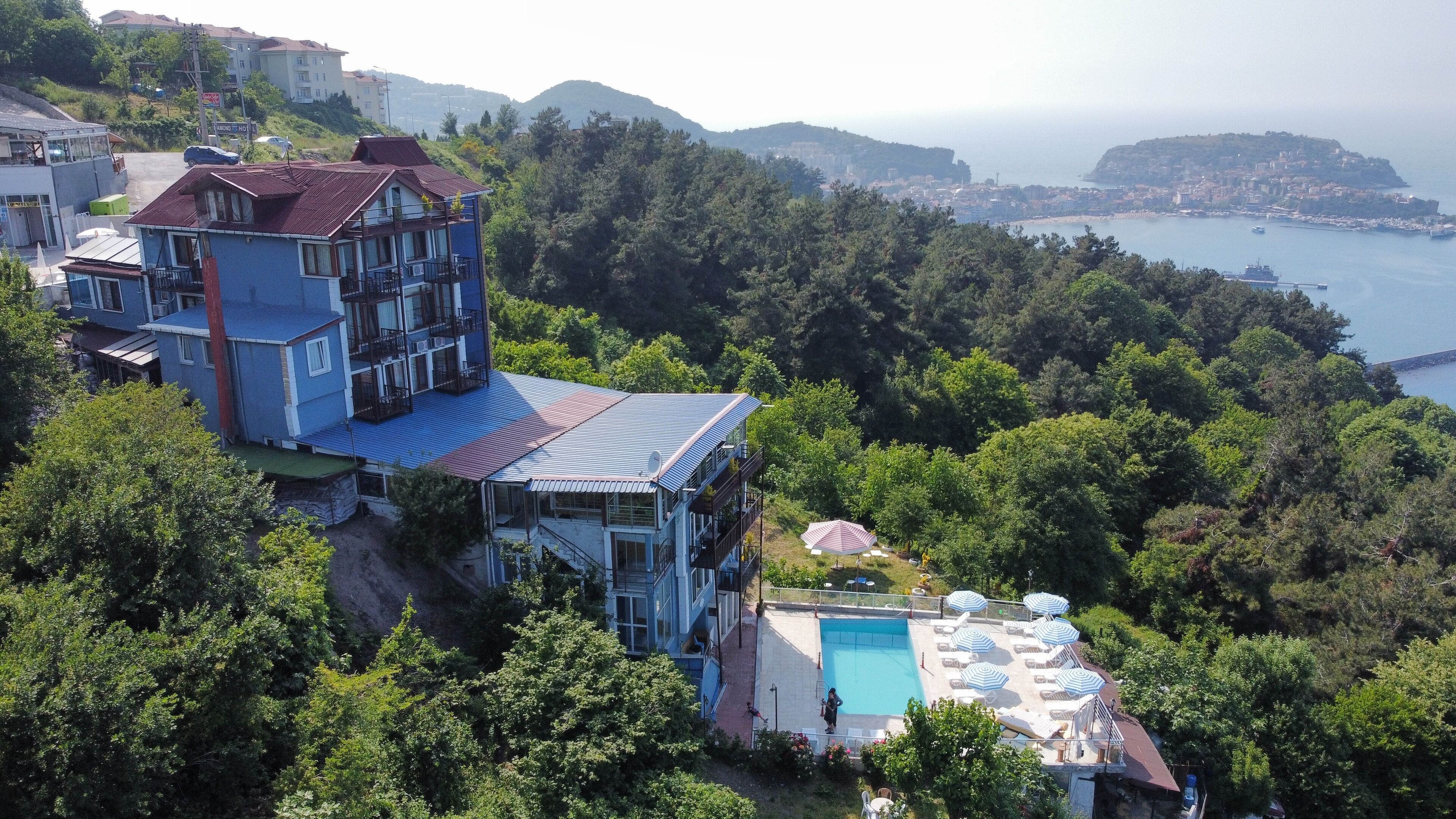 Um edifício com vários andares, piscina no terraço e área de estar ao ar livre, rodeado por vegetação densa e com vista para uma cidade costeira.