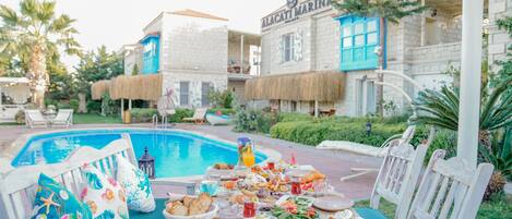 Disposição para refeições à beira da piscina, com mesa, cadeiras e buffet de comida e bebidas.