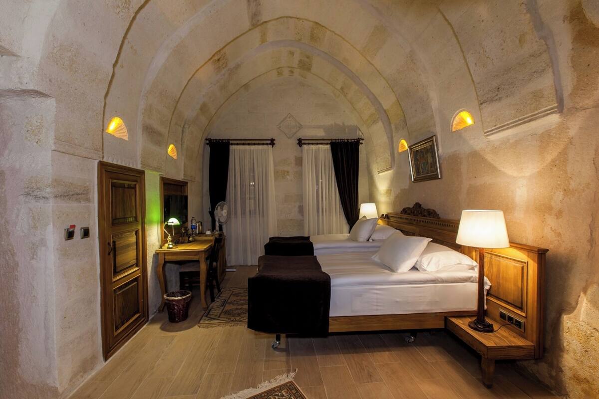 Quarto com uma cama grande, uma escrivaninha com abajur e uma cadeira. Há uma janela com cortinas, e as paredes têm teto em arco.