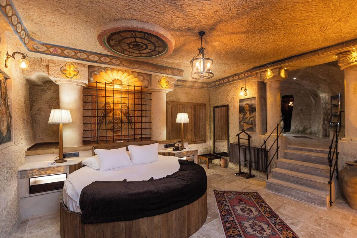 Um quarto luxuoso com uma cama grande, cabeceira de madeira e teto decorativo. Existe uma escada que leva a um andar superior.