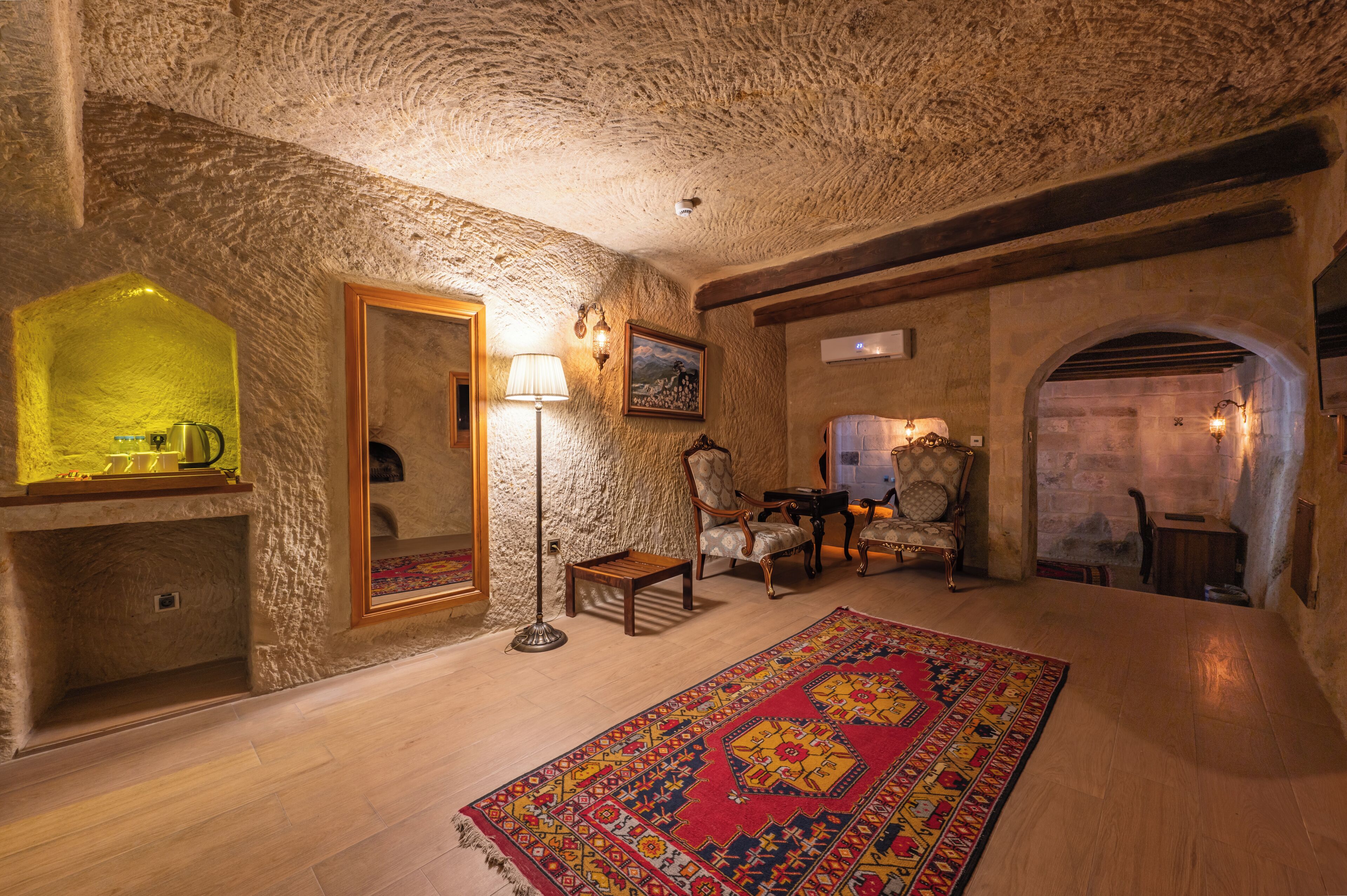 Quarto rústico com paredes de pedra, piso de madeira, tapete estampado, lareira e lustre.
