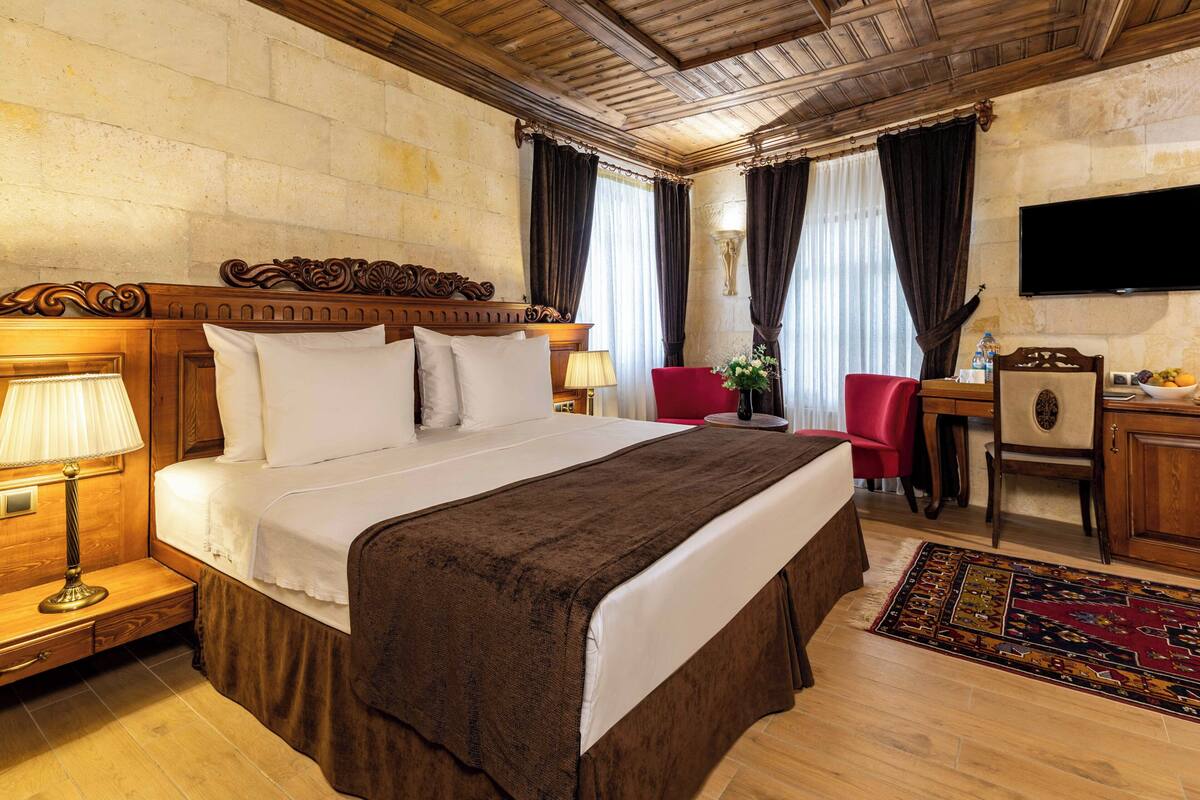 Quarto de hotel com cabeceira de madeira, cama grande, escrivaninha com cadeira, televisão e tapete estampado.