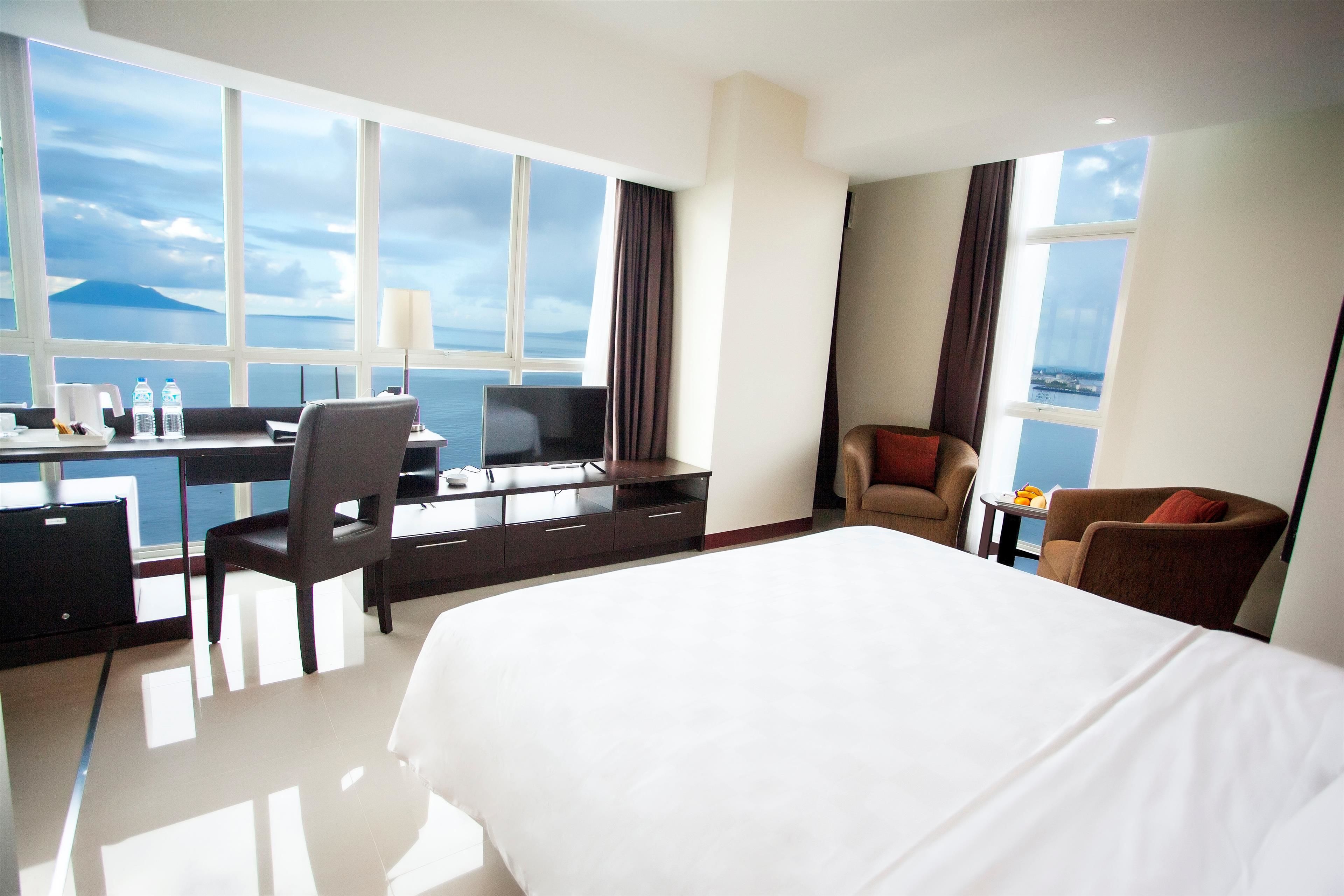 Quarto de hotel com uma cama grande, uma escrivaninha com cadeira, televisão e vista para o mar.