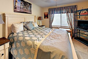 Quarto com cama, mesas de cabeceira, luminária, prateleira com objetos decorativos e janela com cortinas.