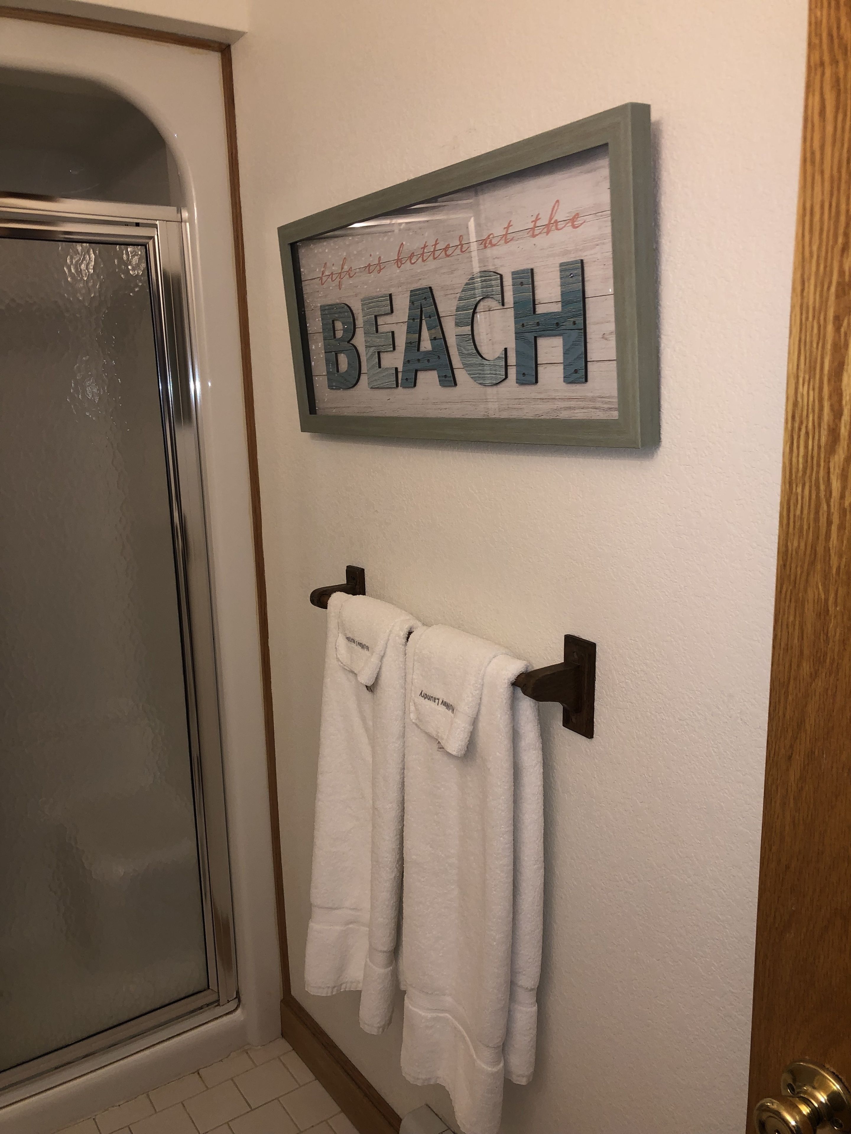 Um banheiro com divisória de box de vidro, um porta-toalhas com duas toalhas brancas e uma placa emoldurada acima do porta-toalhas com a inscrição 'A vida é melhor na praia'.
