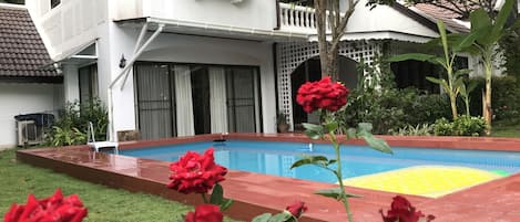 Uma casa branca com piscina, rodeada por vegetação exuberante e rosas vermelhas.