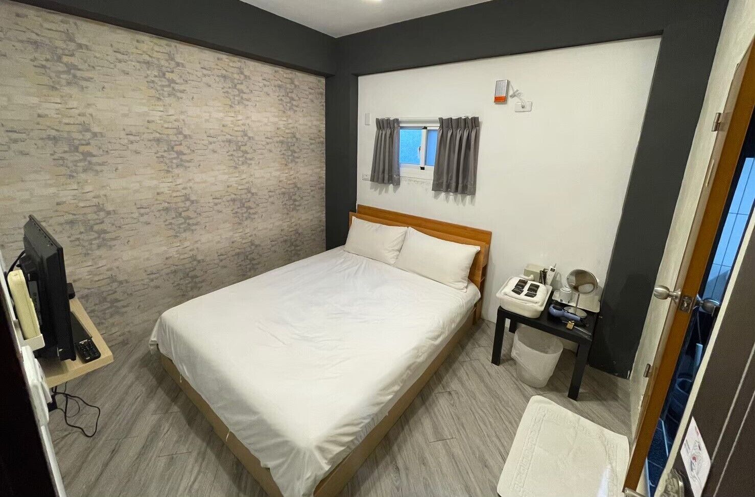 Quarto de hotel com cama, televisão, mesa de cabeceira com telefone e luminária, e janela com cortinas.