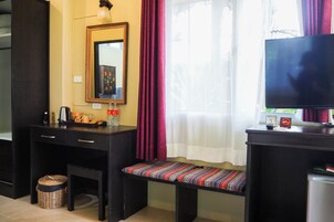 Quarto de hotel com uma escrivaninha de madeira escura, um banco com almofada estampada, uma televisão e uma janela com cortinas.