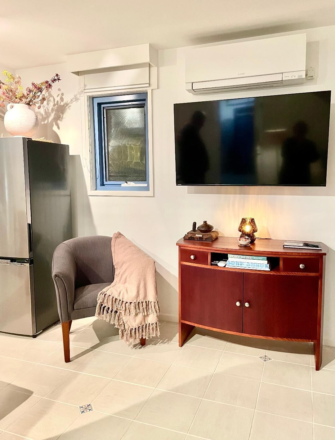Uma sala de estar moderna com uma TV de tela plana na parede, um sofá cinza com manta felpuda, um armário de madeira com livros e uma geladeira de aço inoxidável.