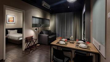 Quarto de hotel moderno com cama, um pequeno sofá, uma mesa de jantar posta para dois e uma janela grande com cortinas.