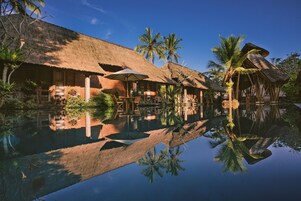 Um resort com construções de telhado de palha, uma piscina refletindo as estruturas e palmeiras.