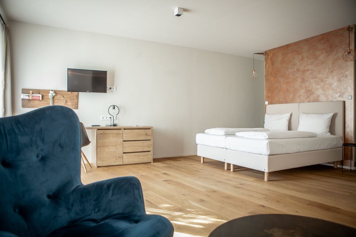 Quarto moderno com chão de madeira, cama branca, cadeira azul, cómoda de madeira e televisão na parede.