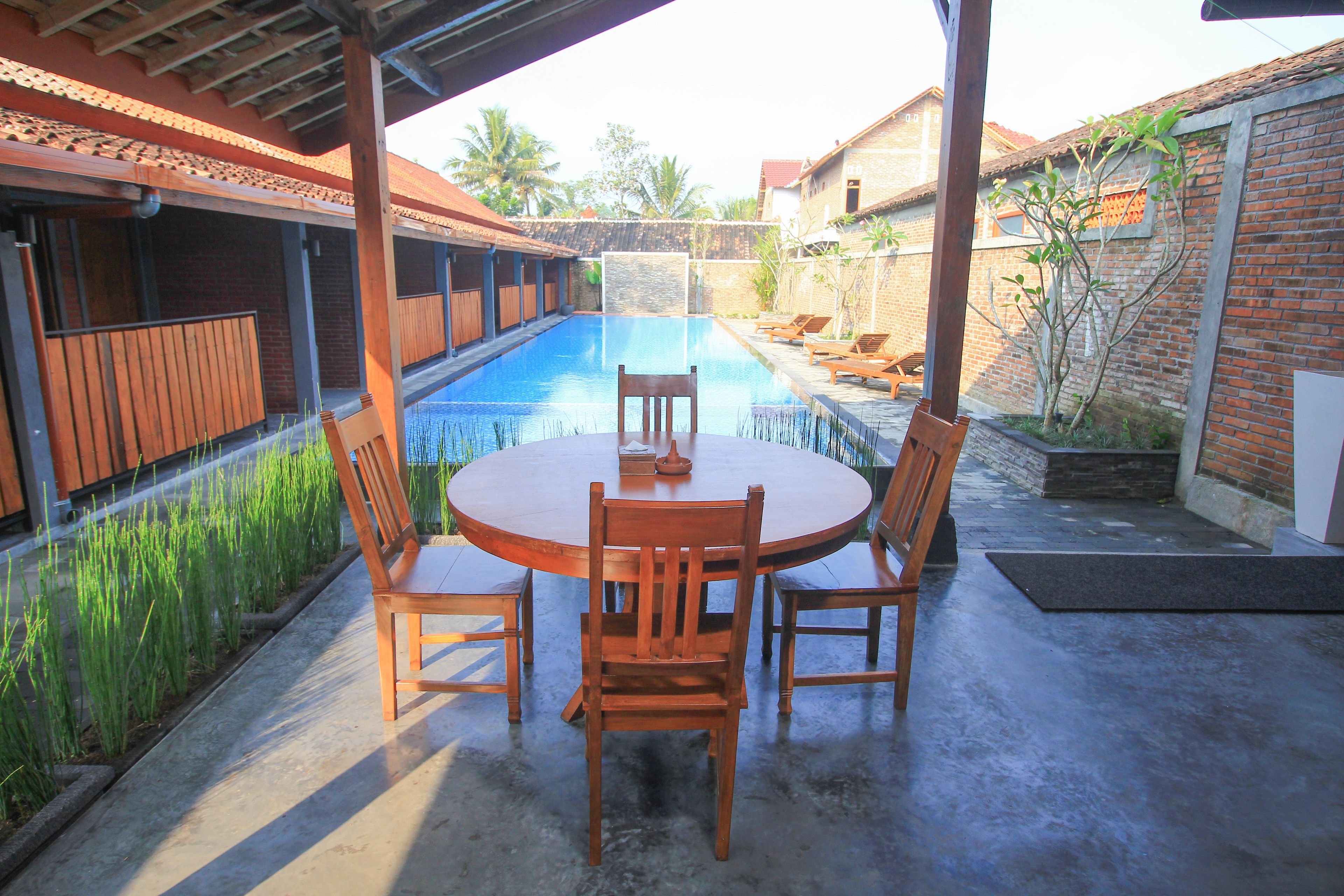Mesa de madeira redonda com quatro cadeiras, piscina e área de varanda coberta com móveis de madeira e plantas.