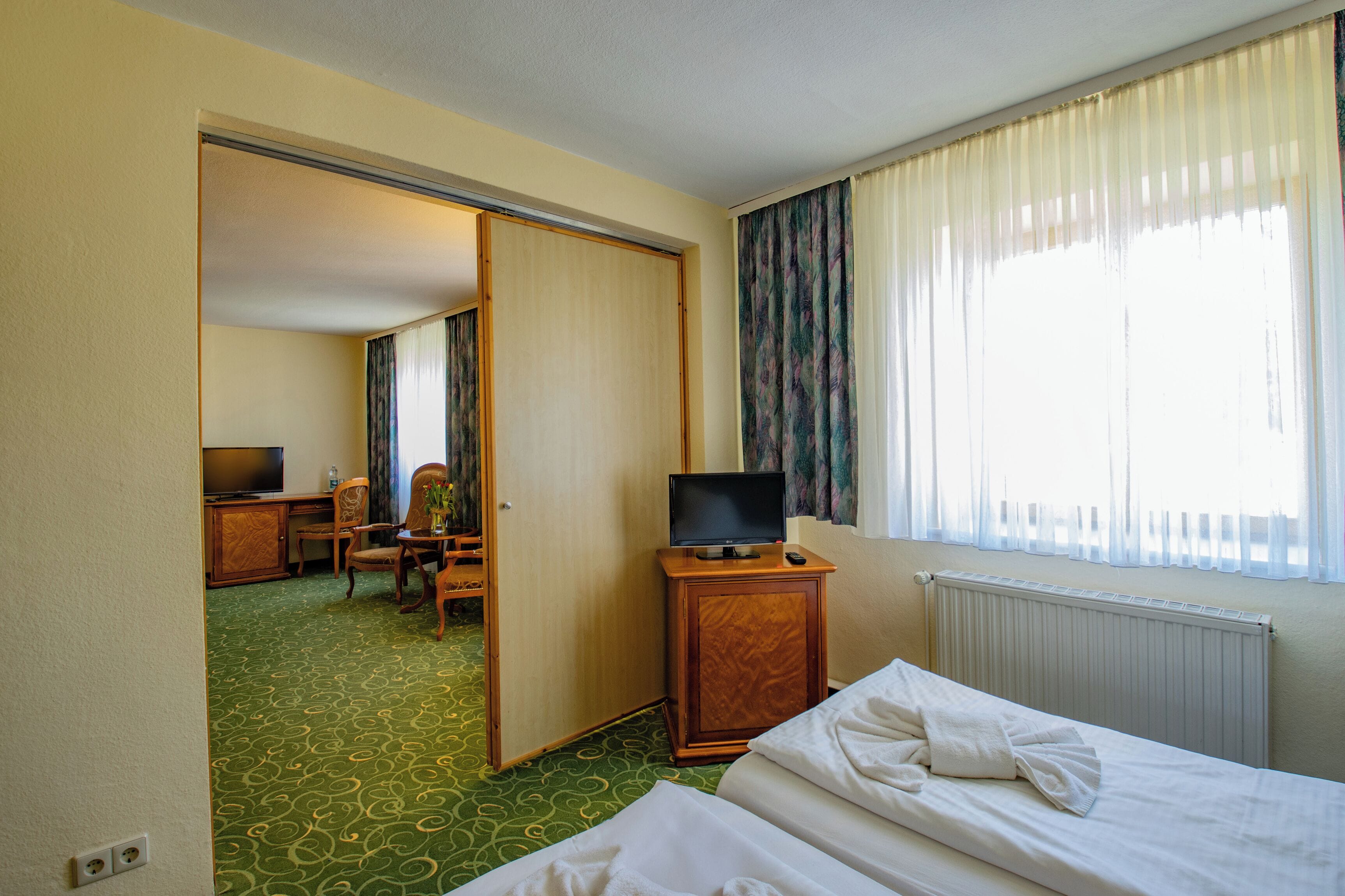 Quarto de hotel com cama, escrivaninha, televisão, cadeira e janela com cortinas.