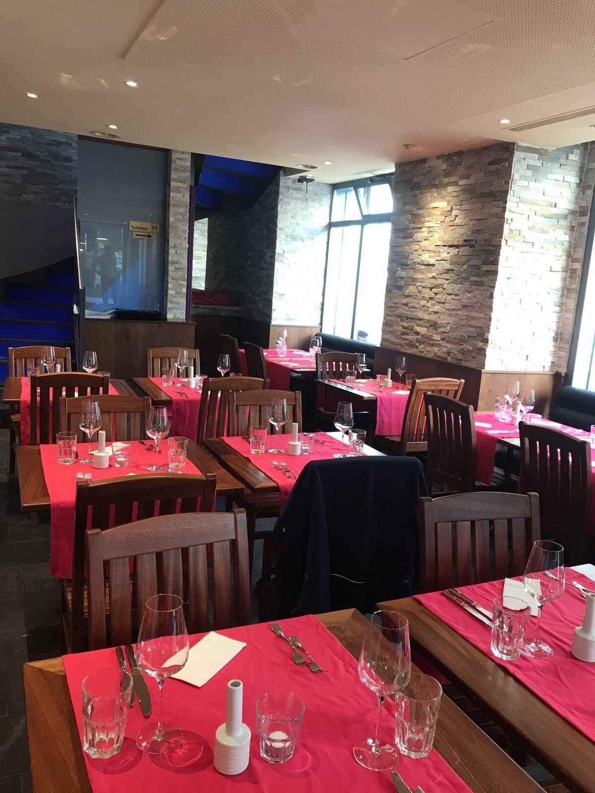 Um restaurante com várias mesas dispostas, cobertas por toalhas vermelhas, com taças de vinho e talheres. O interior apresenta paredes de tijolos aparentes e grandes janelas que permitem a entrada de luz natural.