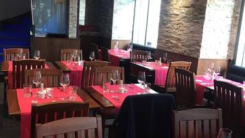 Um restaurante com várias mesas dispostas, cobertas por toalhas vermelhas, com taças de vinho e talheres. O interior apresenta paredes de tijolos aparentes e grandes janelas que permitem a entrada de luz natural.