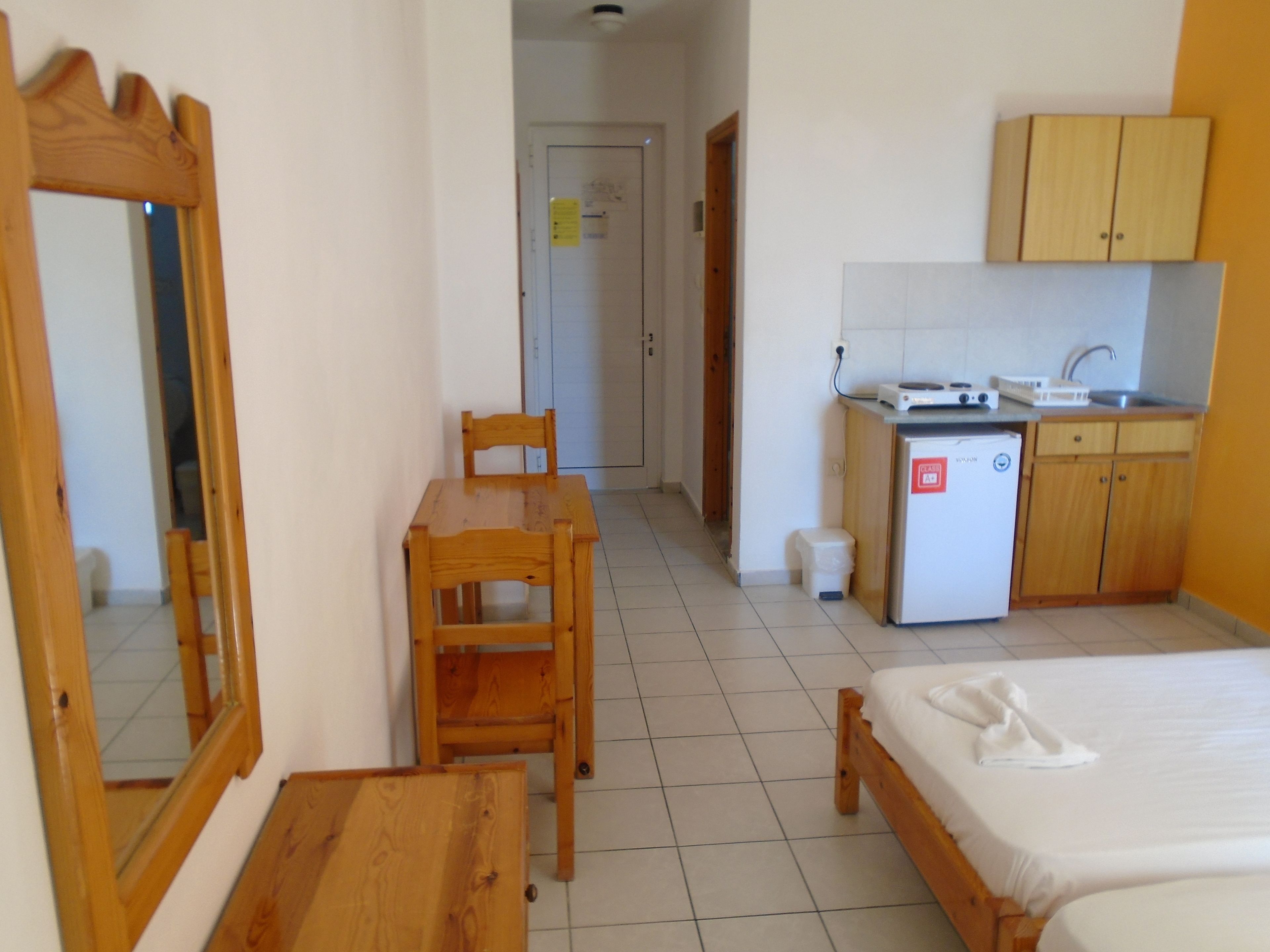 Quarto de hotel compacto com cama, escrivaninha de madeira, cadeira, pequeno espaço de cozinha e banheiro.