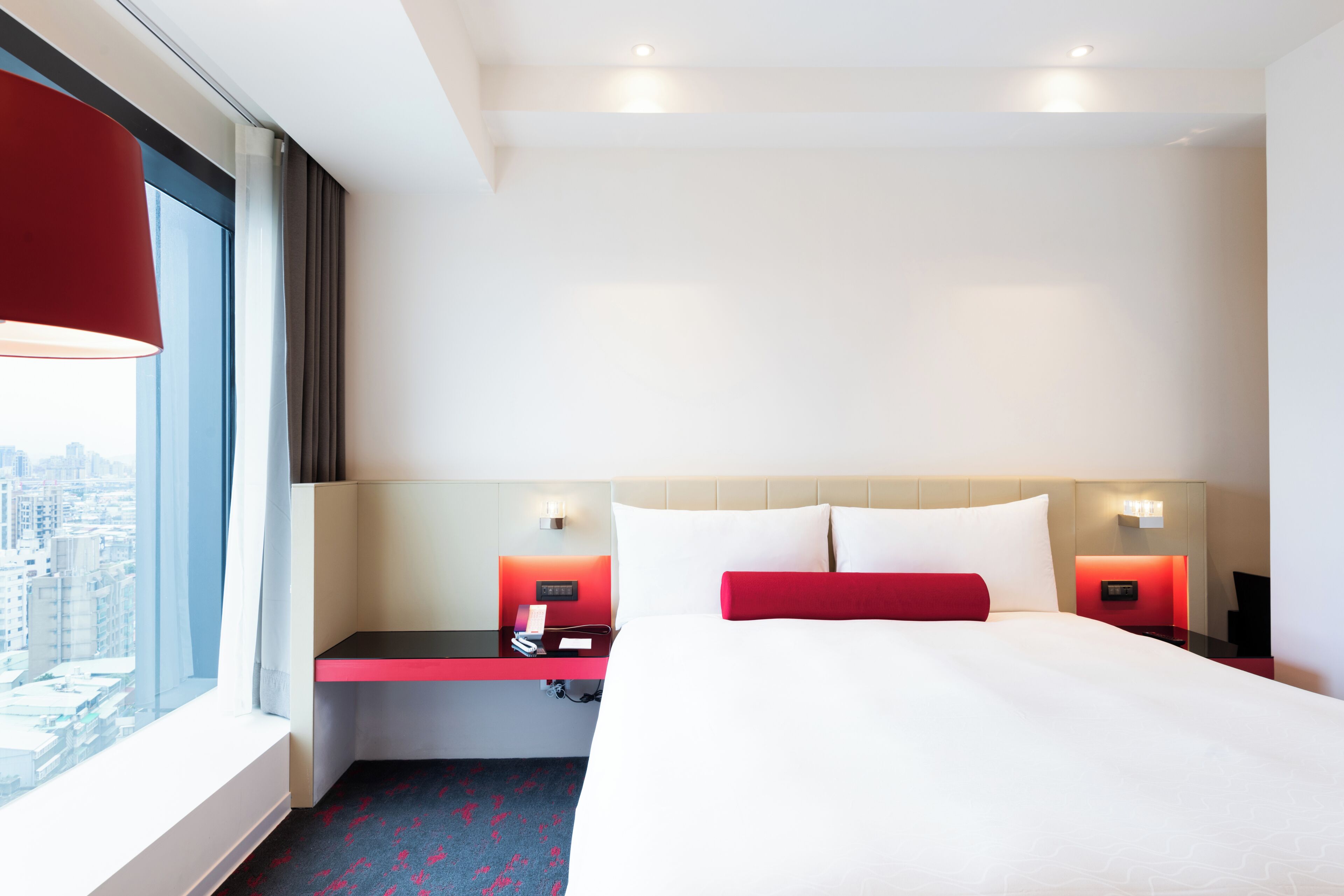 Quarto de hotel com cama, mesas de cabeceira, escrivaninha e janela com vista para a cidade.