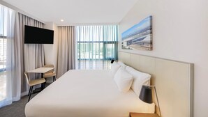 Quarto de hotel com uma cama grande, uma mesa, uma cadeira, televisão na parede e janela com cortinas.