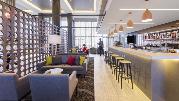 Um lobby moderno de hotel com área de bar, disposição de assentos e uma grande janela que proporciona luz natural.