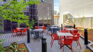Um pátio externo com mesas e cadeiras, cercado por vegetação e edifícios modernos.