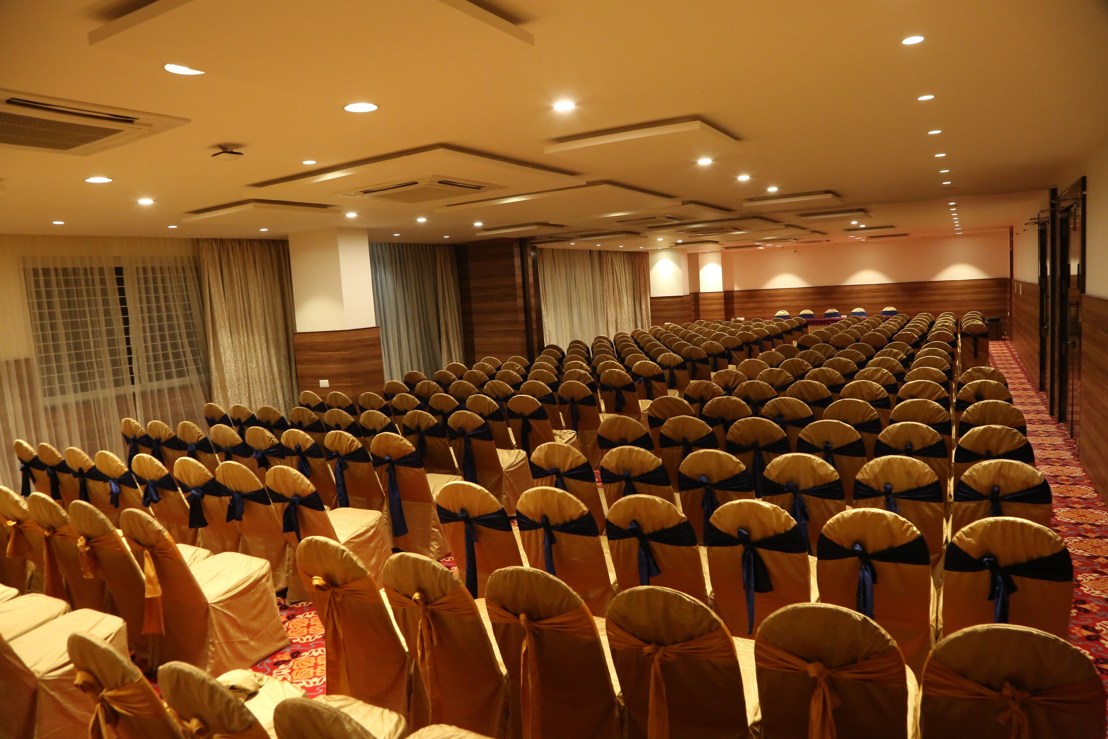Uma sala de conferências com fileiras de cadeiras adornadas com fitas bege e azuis, painéis de madeira nas paredes e piso coberto de carpete.