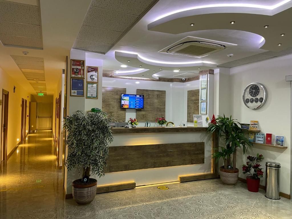 Uma área de recepção de hotel, com balcão, tela digital e relógio na parede.