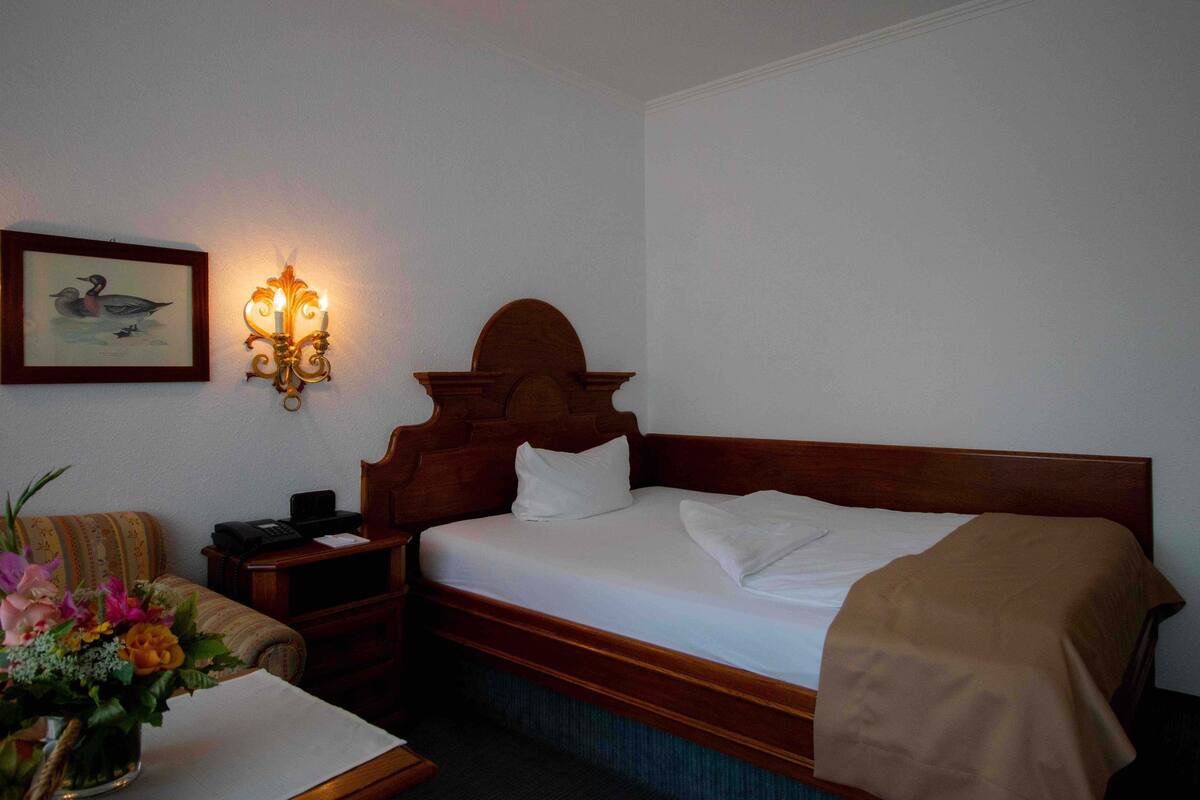 Quarto de hotel com cama de madeira, mesa de cabeceira com luminária, quadro na parede e arranjo floral sobre uma mesa.