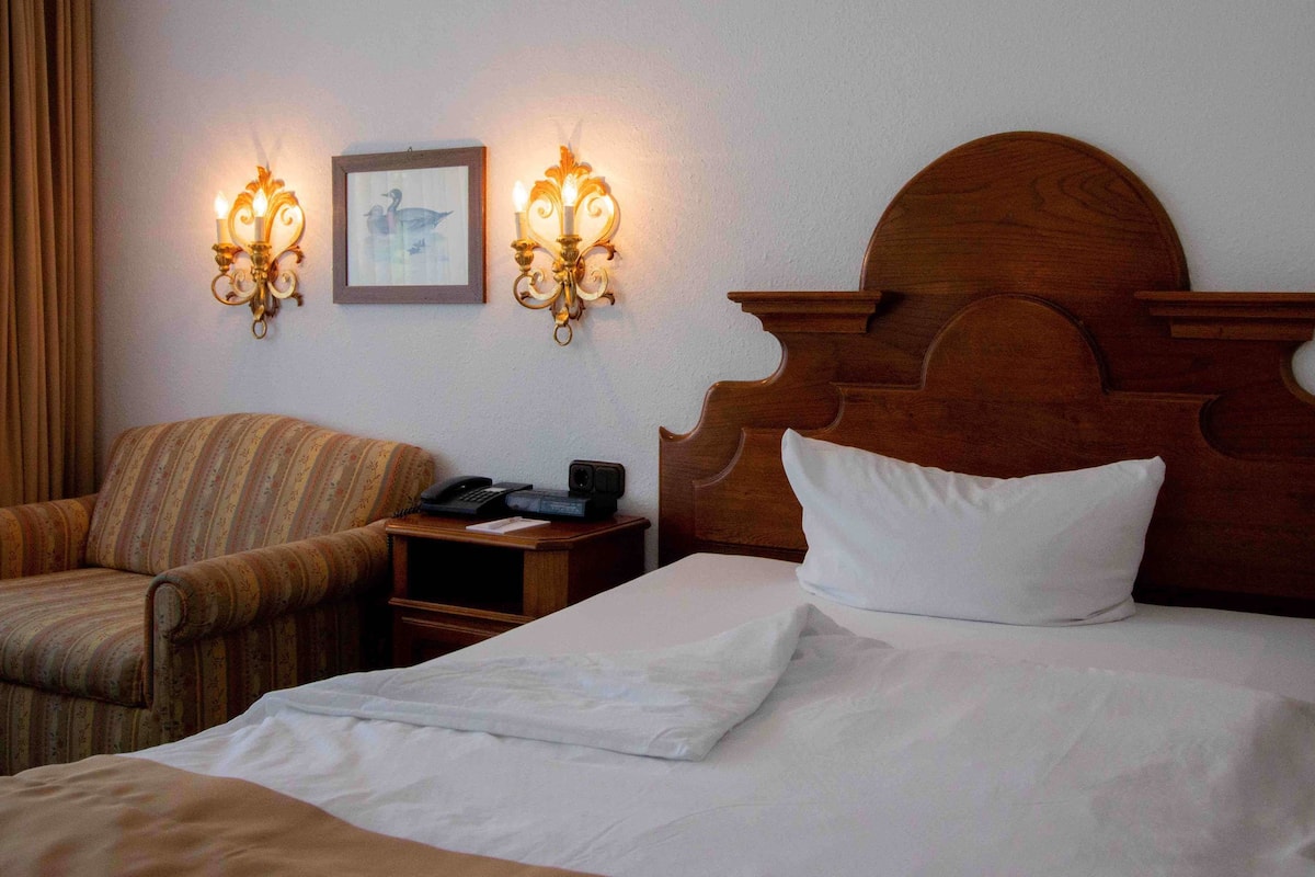 Quarto de hotel com uma cama grande, cabeceira de madeira, mesa de cabeceira com telefone e duas luminárias na parede.