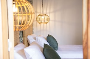 Uma cama com almofadas brancas e verdes, uma luminária de fibra natural e uma parede com textura.