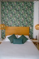 Cama bem arrumada com lençóis brancos e almofadas verdes, cabeceira de vime e um padrão de papel de parede tropical.