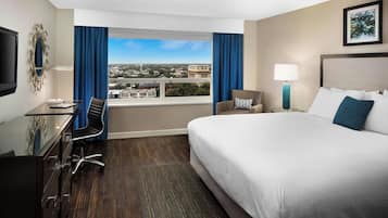 Quarto de hotel com uma cama grande, uma escrivaninha, uma cadeira, televisão e vista para a paisagem urbana.