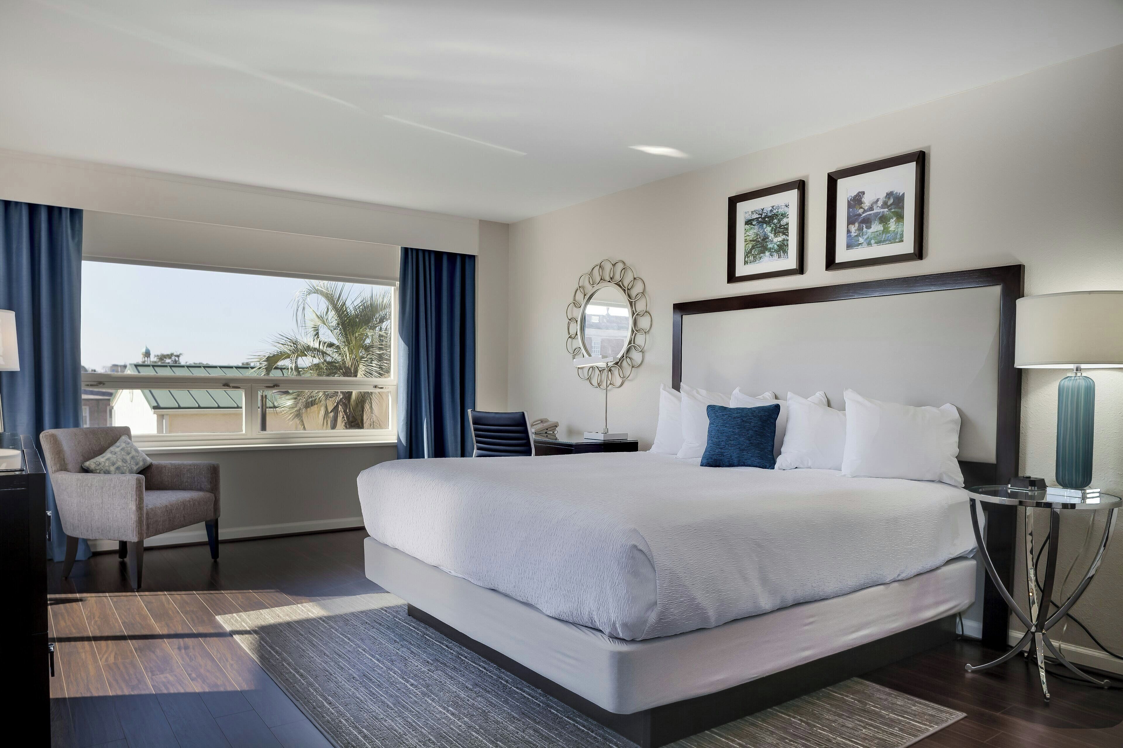 Quarto de hotel com uma cama grande, uma escrivaninha com candeeiro, uma cadeira e vista para o exterior.