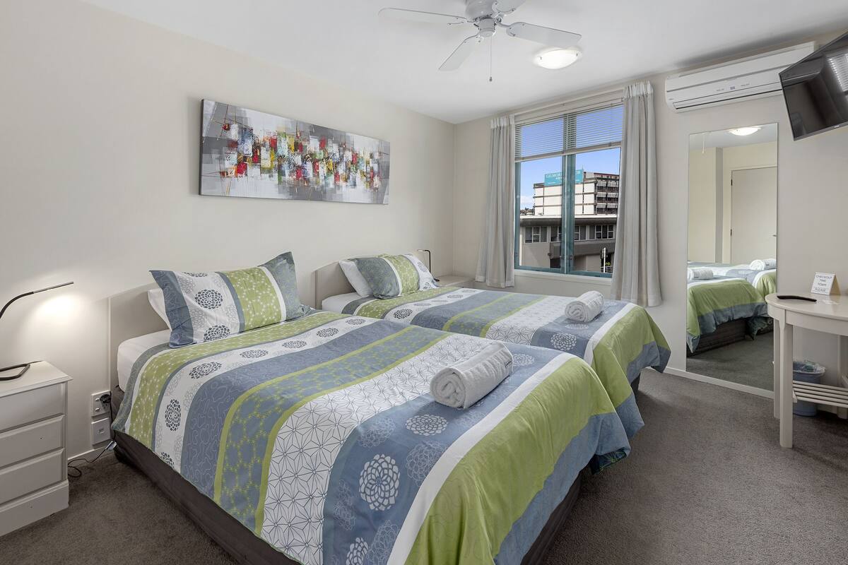 Quarto de hotel com duas camas, um ventilador de teto, uma janela grande com cortinas, uma televisão de tela plana e um quadro na parede.
