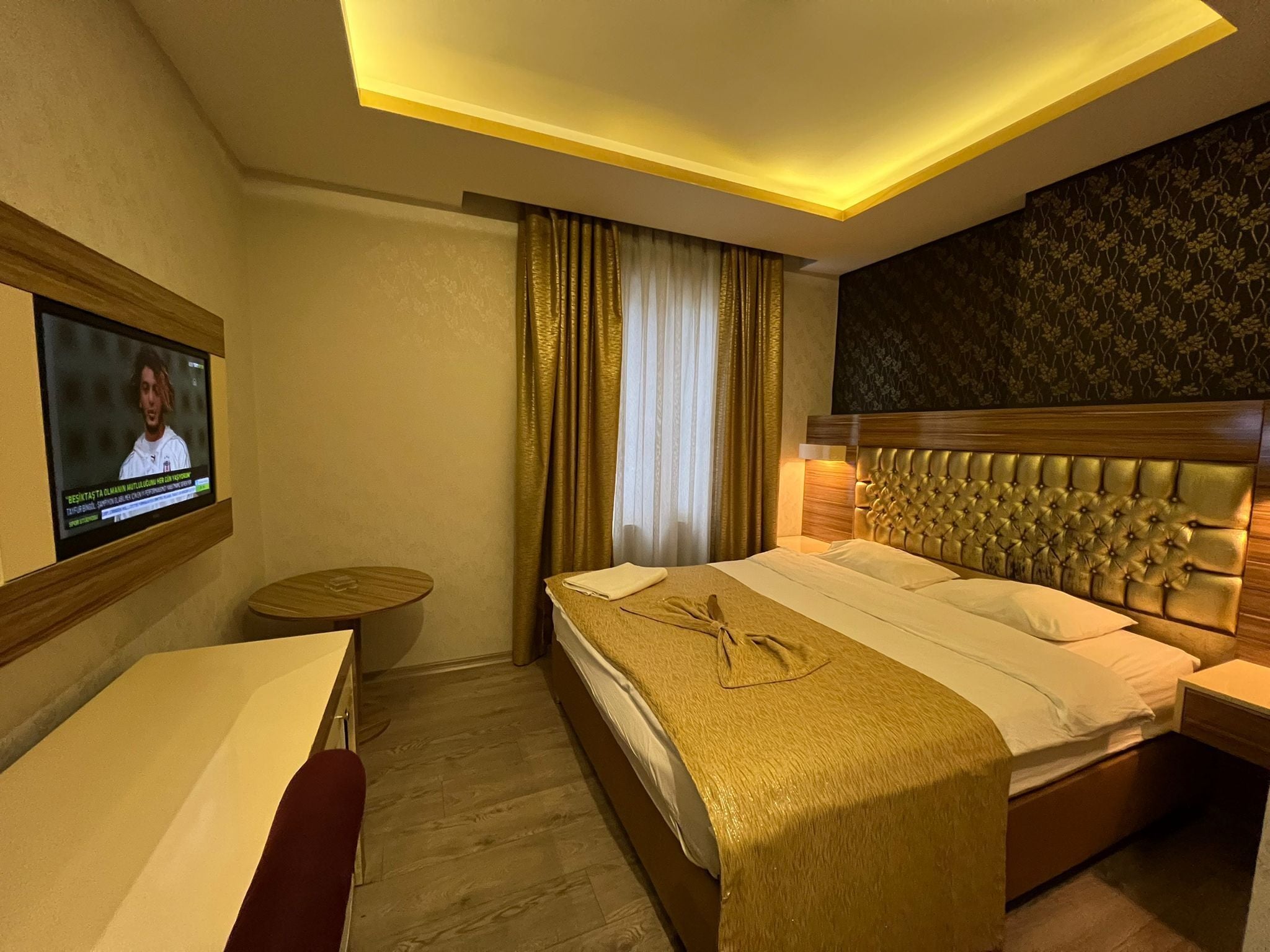 Quarto de hotel com cama, mesa de cabeceira, televisão e cortinas.