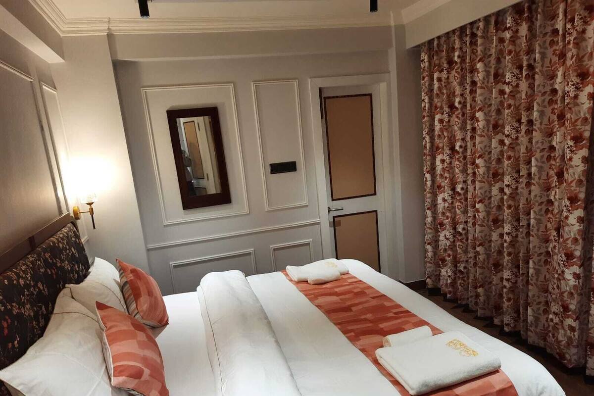 Quarto de hotel com uma cama grande, uma mesa de cabeceira, um espelho e cortinas florais.