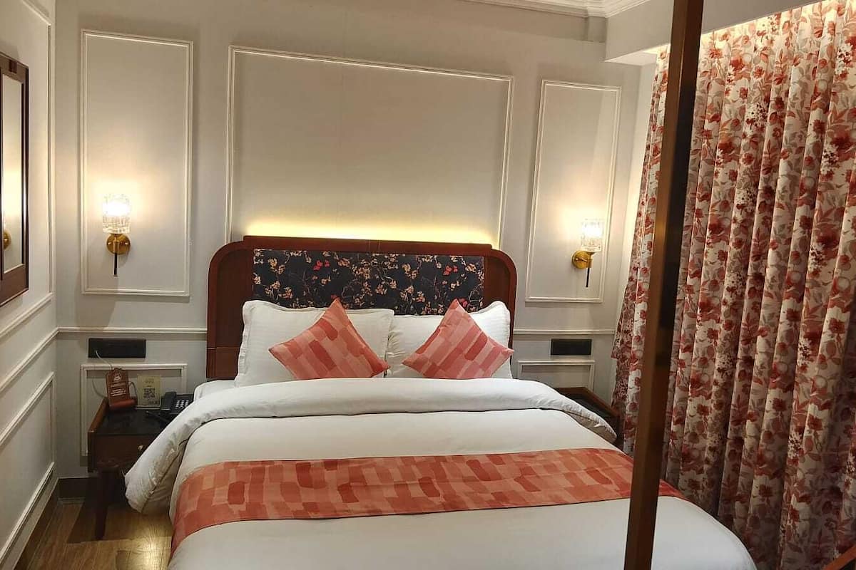 Quarto de hotel com uma cama grande, duas luminárias de cabeceira, uma mesa de apoio e cortina florida.