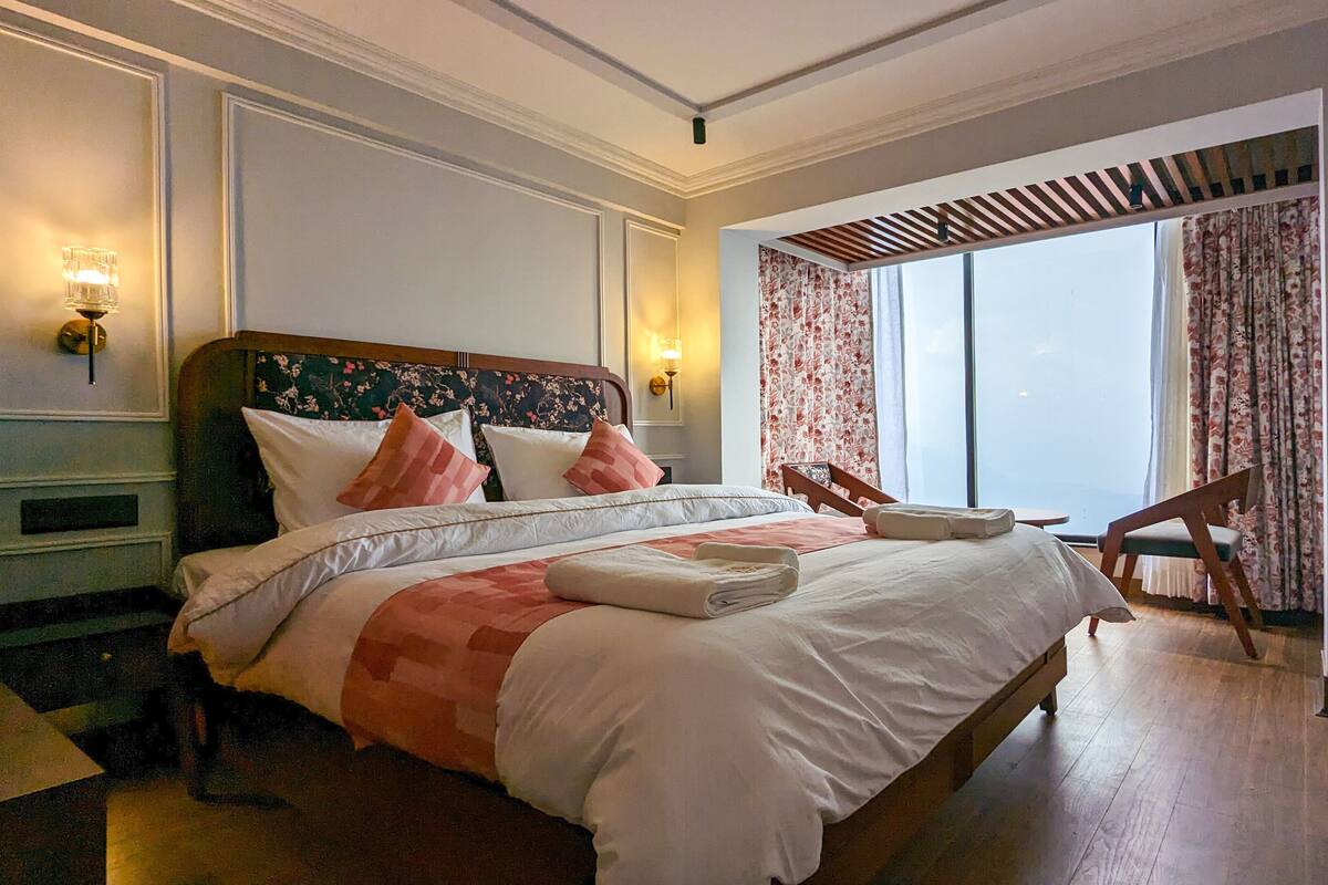 Quarto de hotel com uma cama grande, duas luminárias de cabeceira, um painel de cabeceira de madeira, uma janela com cortinas e uma pequena mesa com uma cadeira.