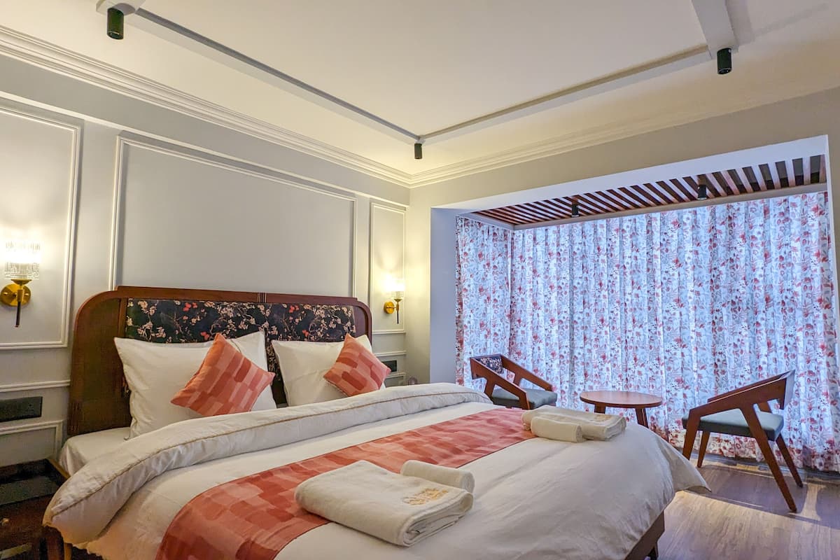 Quarto de hotel com uma cama grande, duas mesas de cabeceira, uma cadeira e uma janela com cortinas florais.