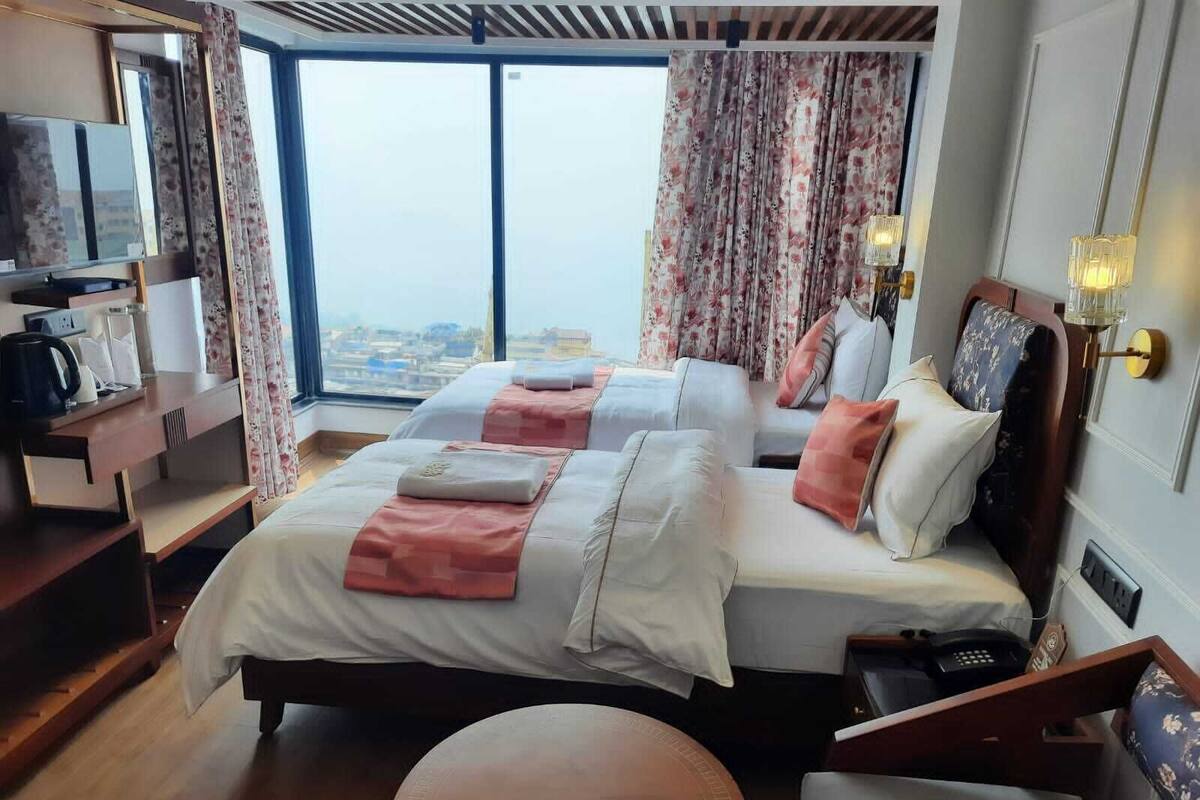 Quarto de hotel com duas camas, uma mesa de escritório com cafeteira, uma cadeira, uma mesa redonda e vista da cidade através de grandes janelas.