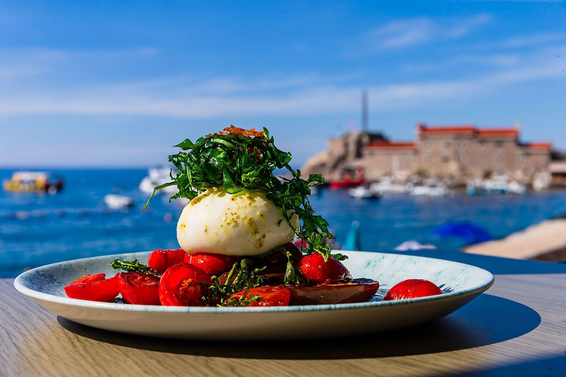Prato de comida com ovo cozido, tomates e vegetais, com um cenário costeiro que inclui barcos e um edifício.
