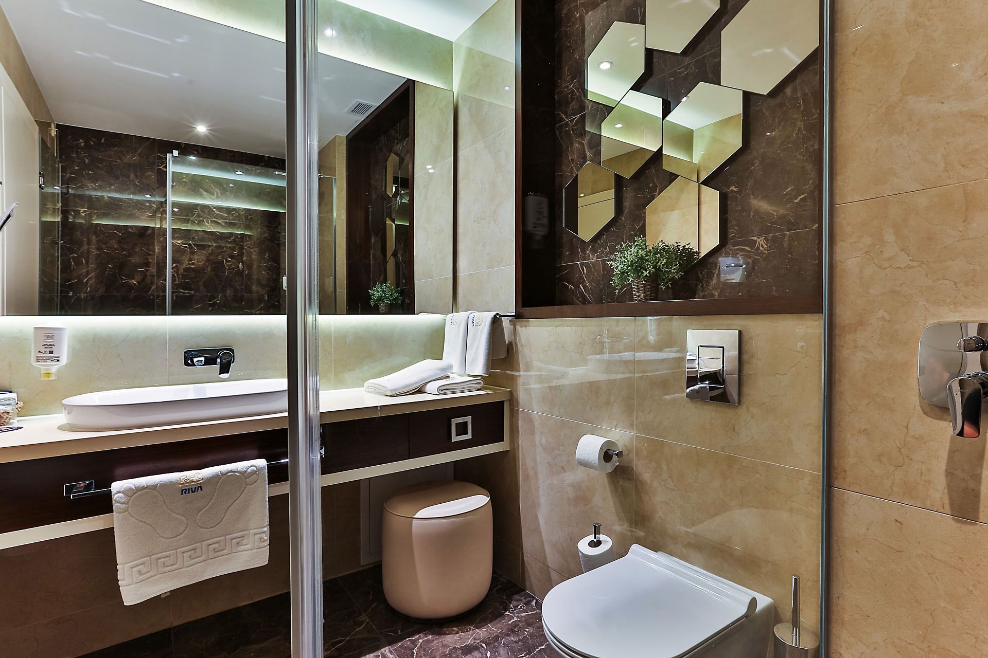 Uma casa de banho moderna com divisória de vidro, lavatório branco, sanita e espelho grande com padrões geométricos.