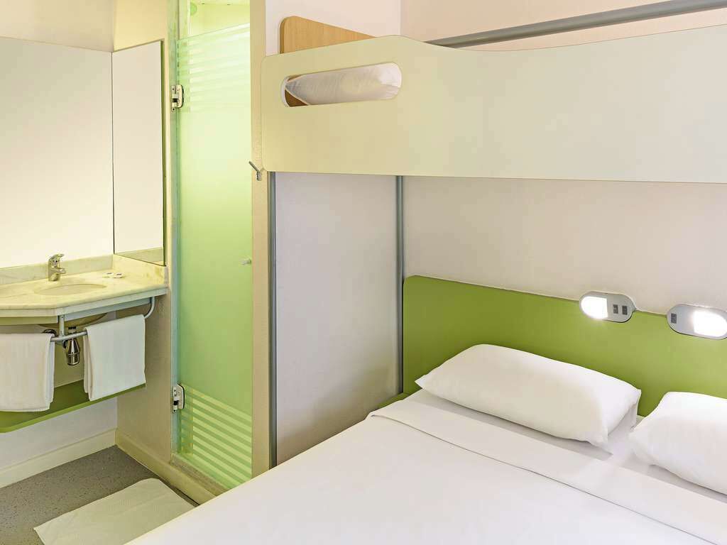 Quarto de hotel com beliche, banheiro com pia e espelho, e cabeceira verde.