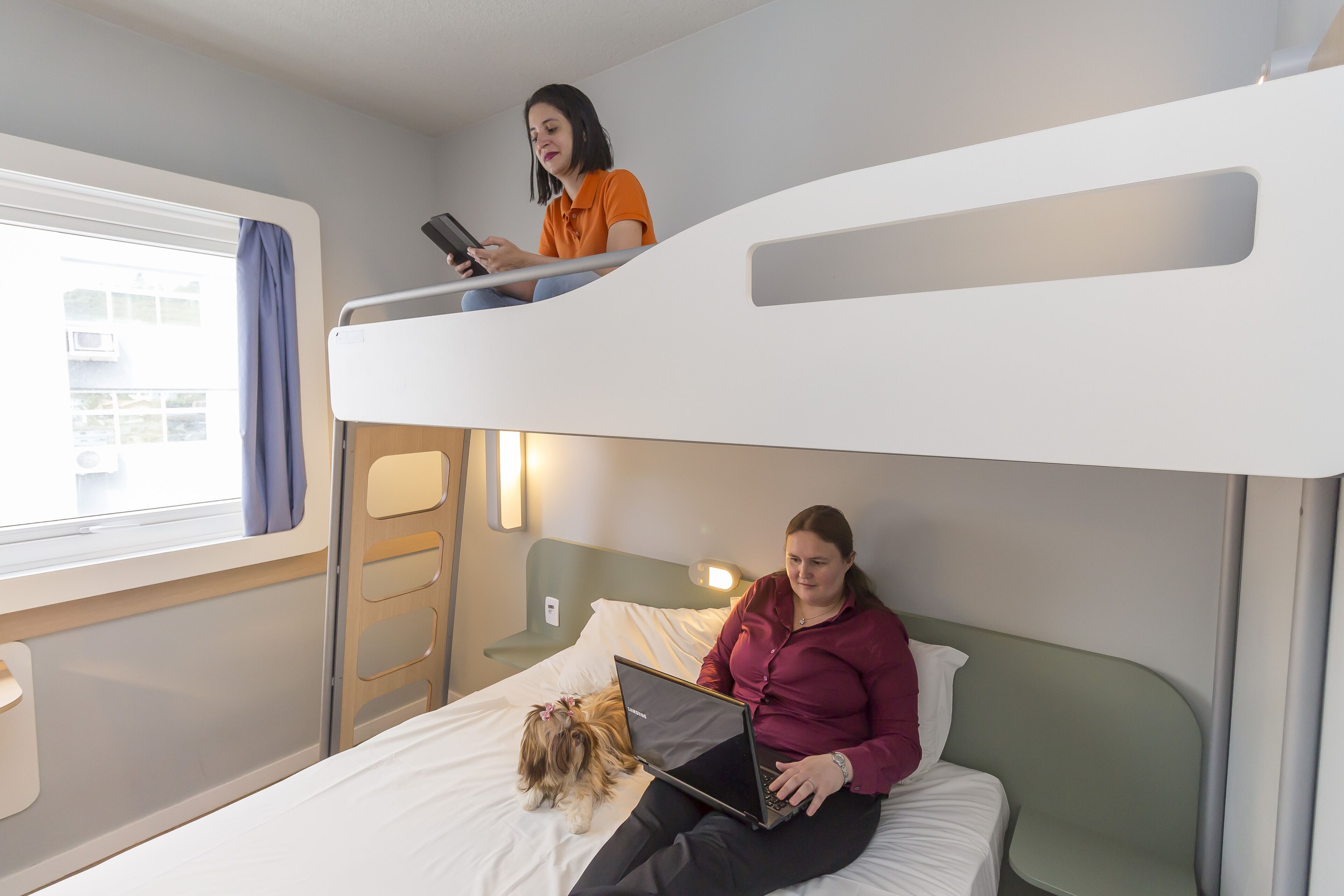 Quarto com beliche, um laptop na cama inferior e uma pessoa sentada na cama superior.