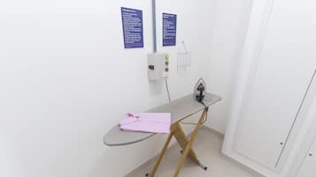 Quarto pequeno com tábua de passar roupa dobrável, ferro e toalha rosa. Há dois avisos informativos na parede.