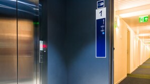Um elevador com visor digital indicando o andar 1, ao lado de um corredor com placa de saída verde e símbolo de acessibilidade para cadeiras de rodas.