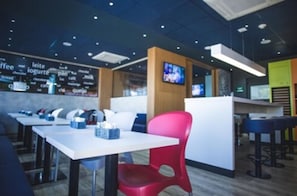 Um café moderno com mesas brancas, cadeiras vermelhas e um menu na parede mostrando diversas opções de comida e bebida.