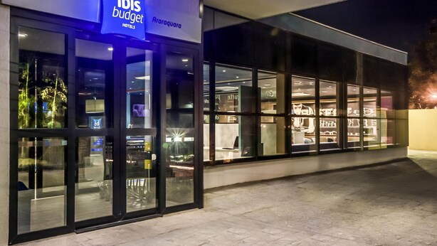Exterior moderno de hotel com portas de vidro, placa com a inscrição 'ibis budget hotels' e interior visível com diversos serviços.