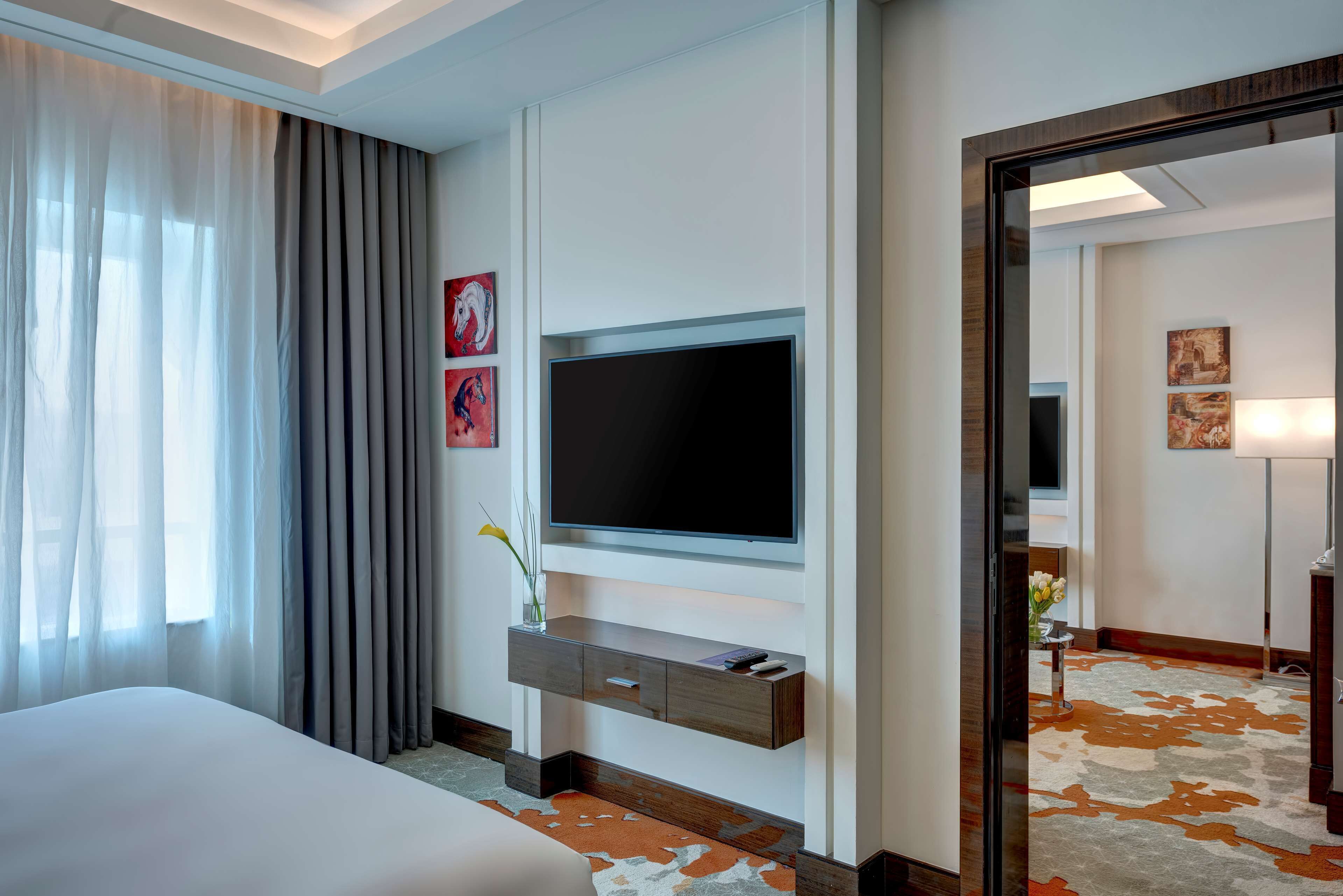 Quarto de hotel com cama, televisão presa na parede, pequena escrivaninha com um vaso e piso coberto por carpete.