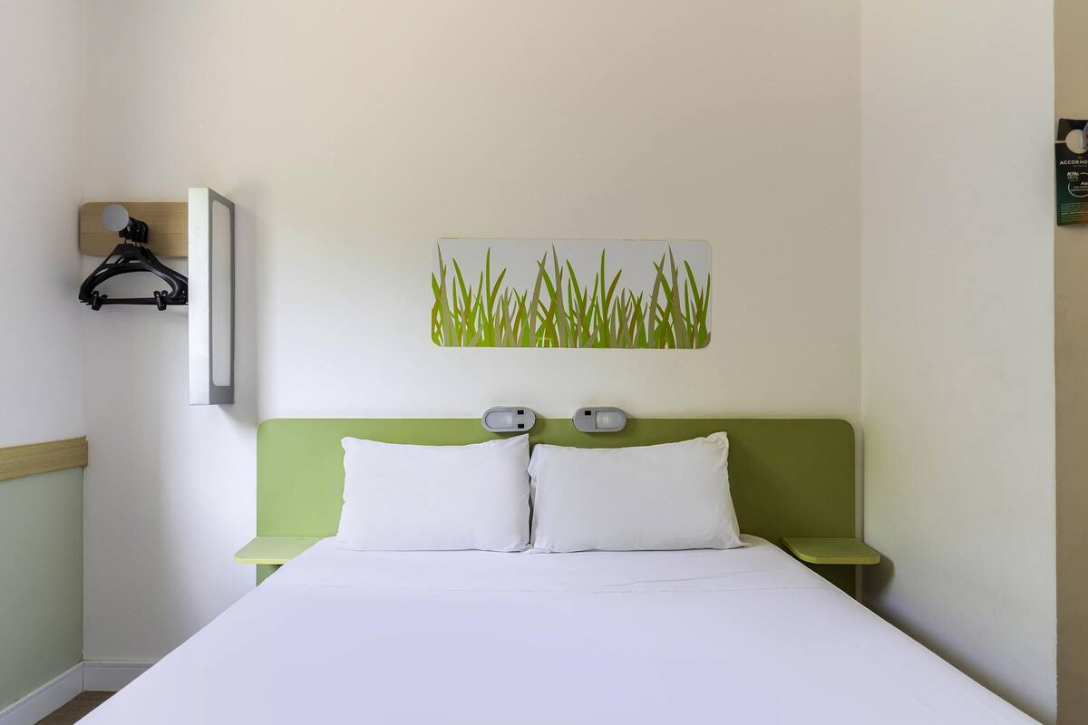 Quarto de hotel com cama, cabeceira verde, almofadas brancas, luminária fixada na parede e arte na parede retratando grama.